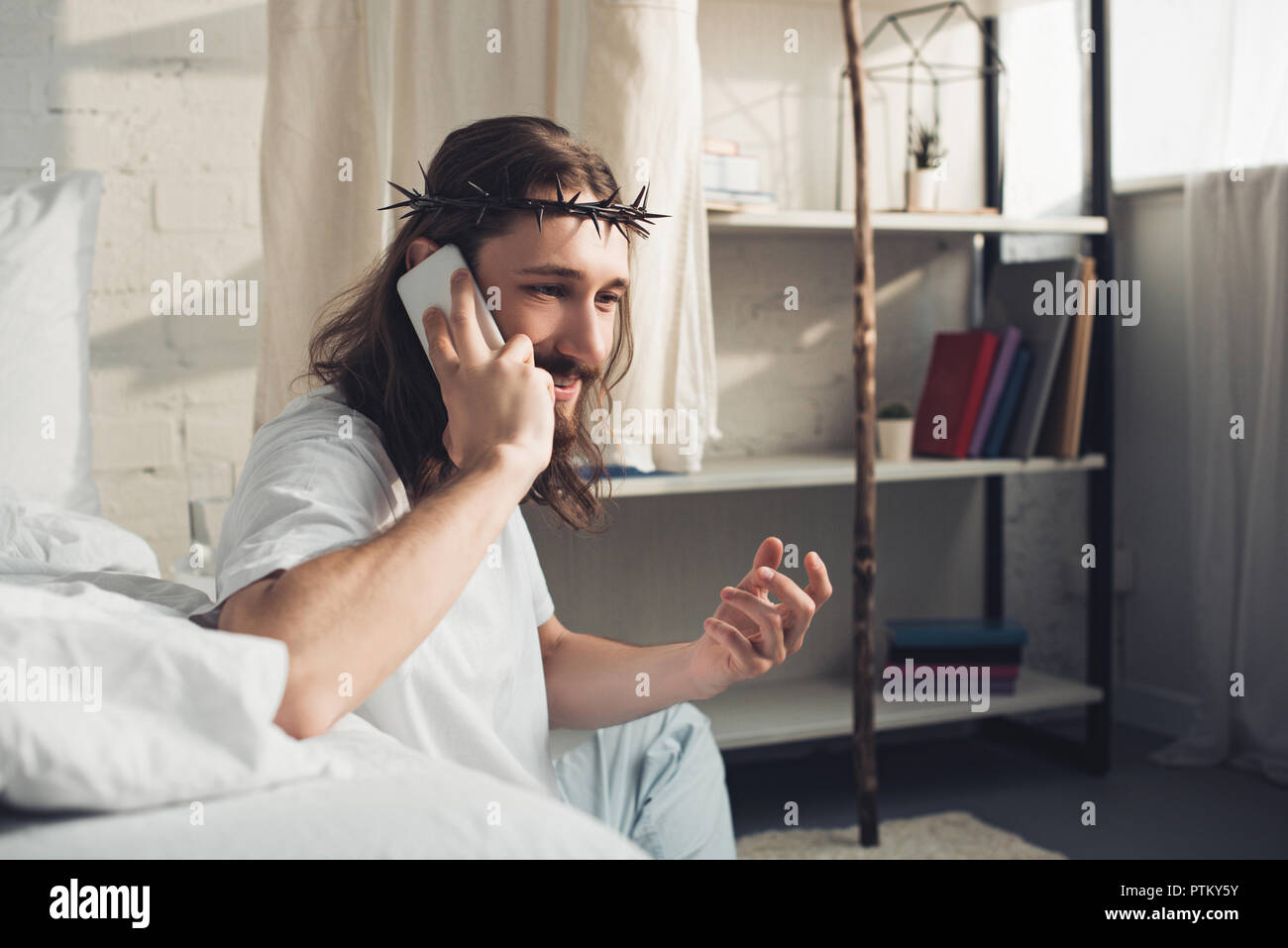 cheerful Jesus in crown of thorns talking on smartphone in bedroom at ...