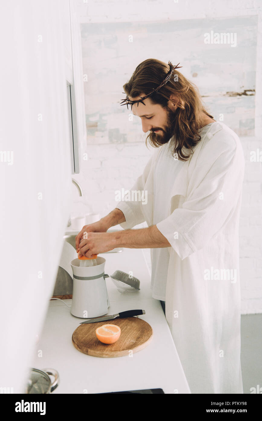 side view of cheerful Jesus doing orange juice by juicer in kitchen at ...