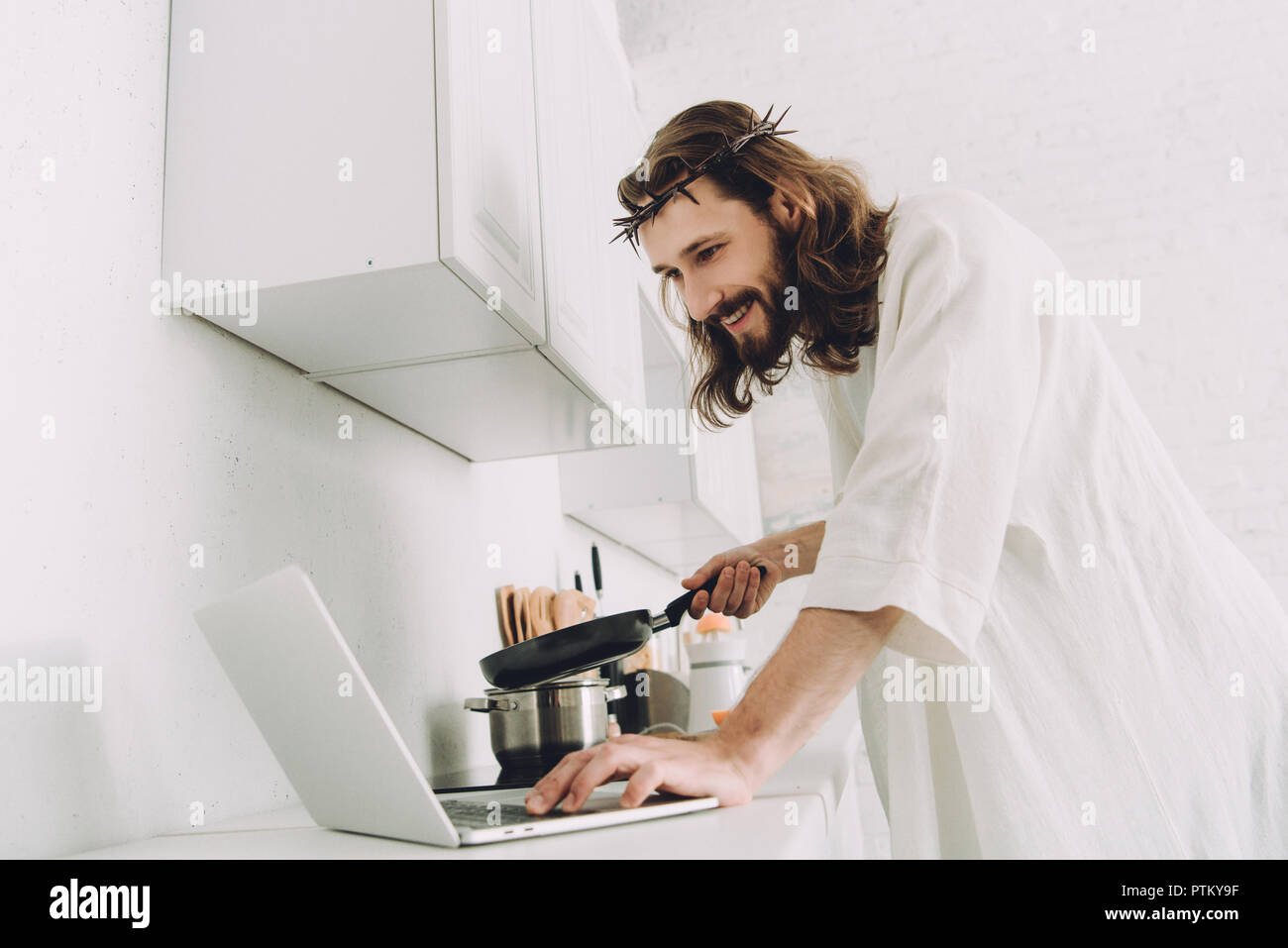 smiling Jesus using laptop while cooking with frying pan in kitchen at ...