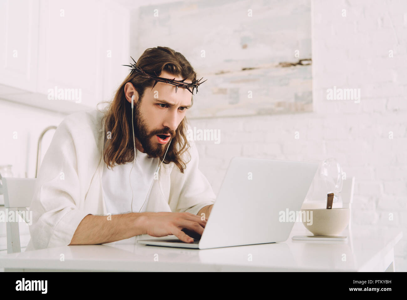 emotional Jesus in crown of thorns and earphones using laptop at table ...