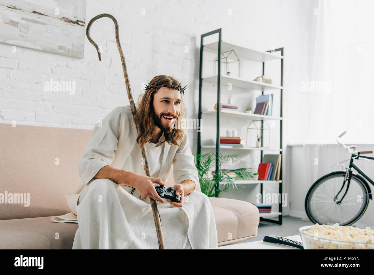 happy Jesus with wooden staff playing video game by joystick on sofa at ...