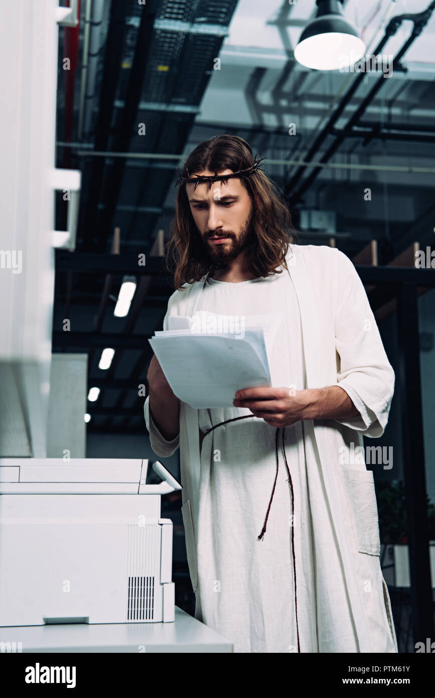 focused Jesus in crown of thorns and robe checking papers near copy ...