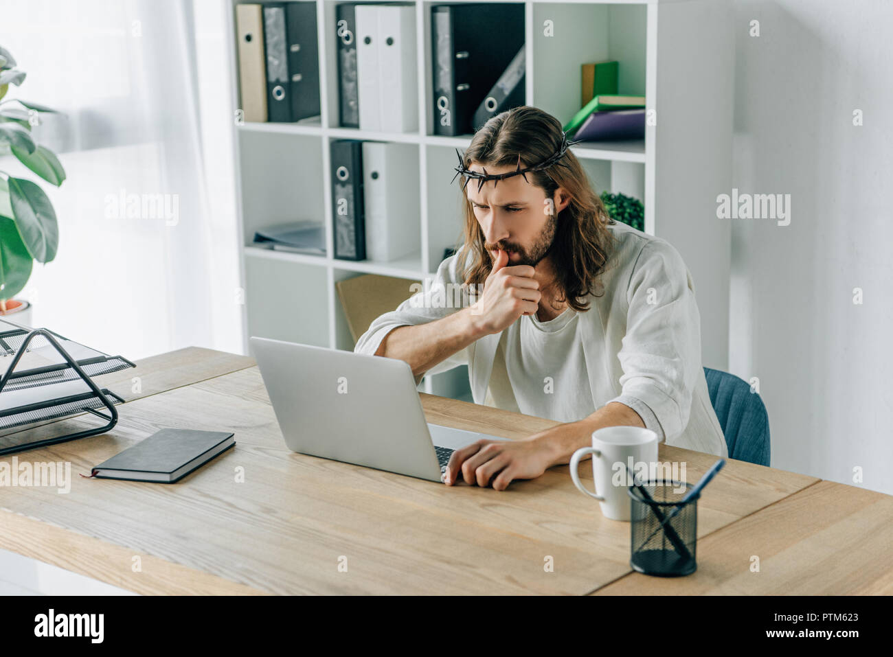 pensive Jesus in crown of thorns using laptop at table in modern office ...