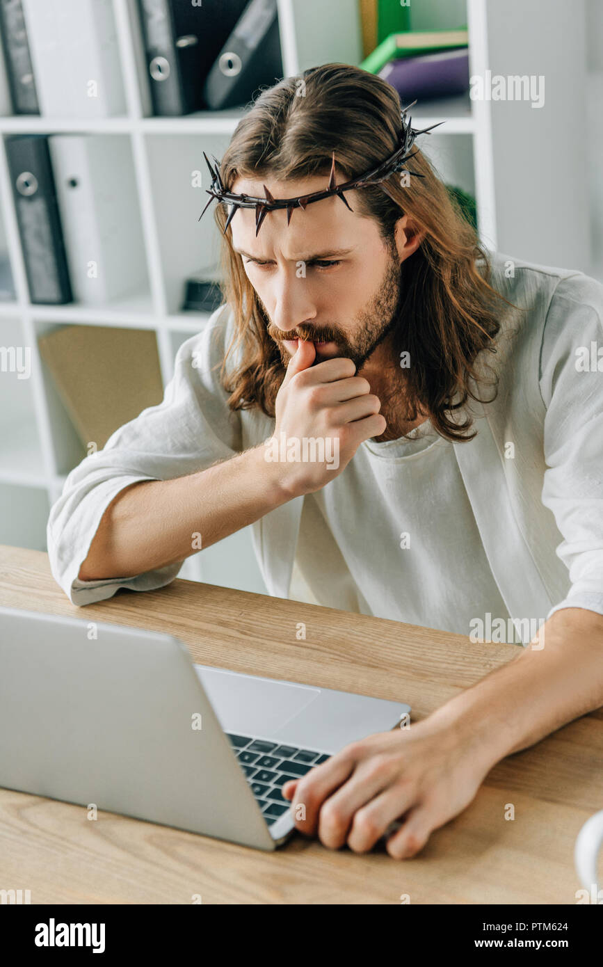 thoughtful Jesus in crown of thorns using laptop at table in modern ...