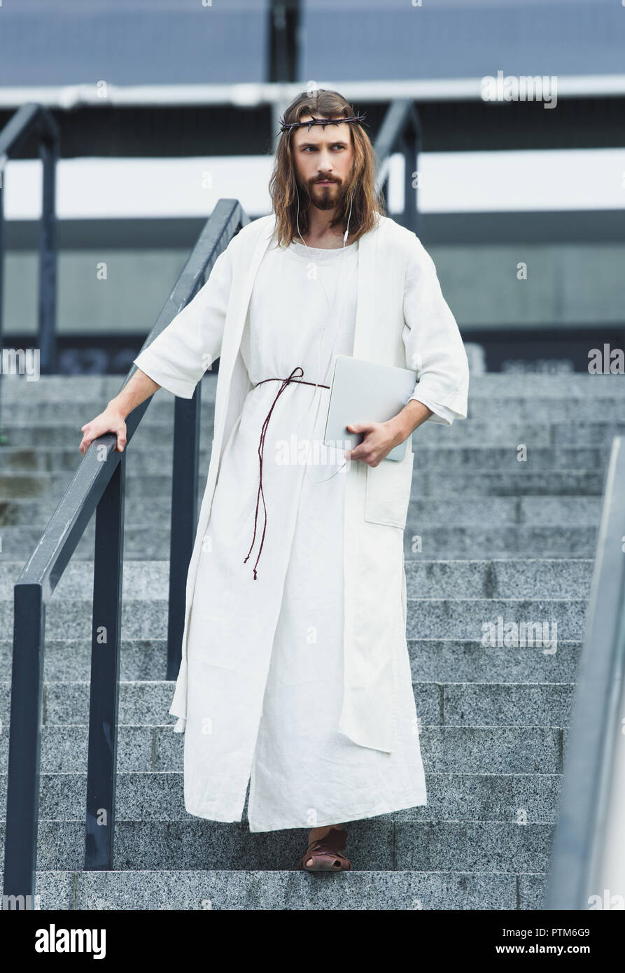 serious Jesus in robe and crown of thorns walking on stairs with laptop ...