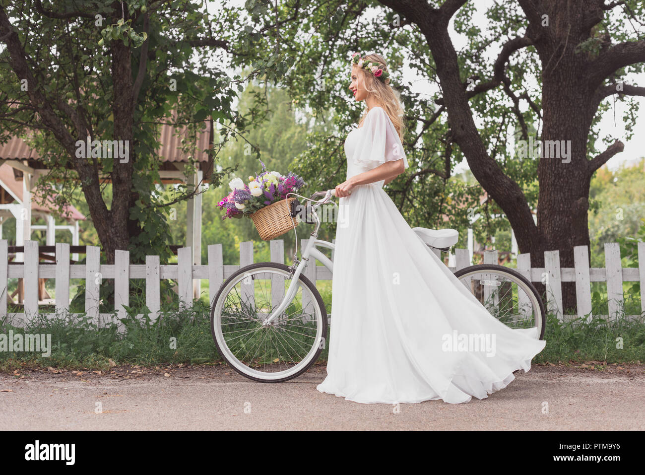 side view of young bride in floral wreath and wedding dress walking ...