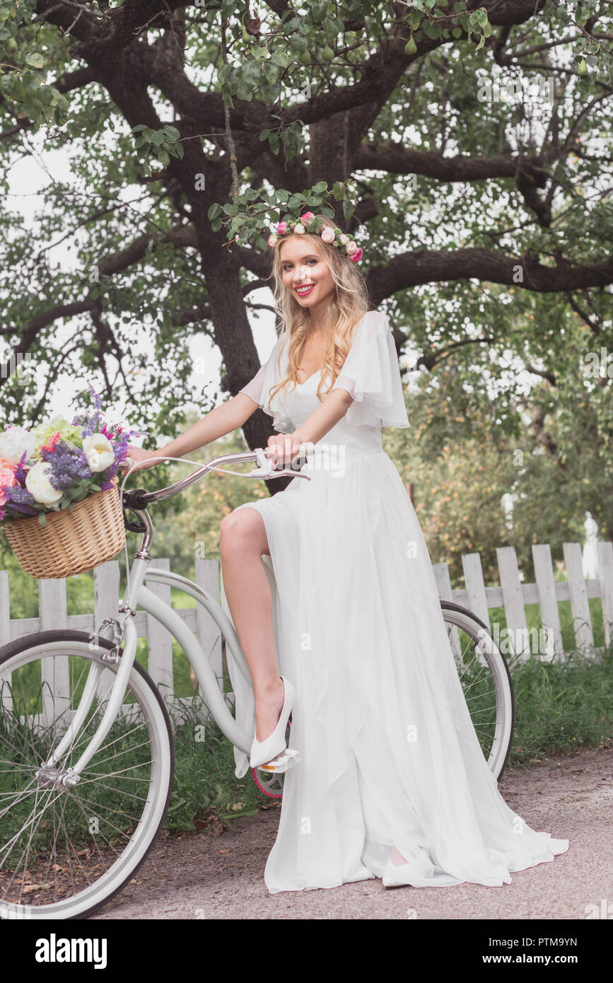 young blonde bride in wedding dress and floral wreath riding bicycle ...