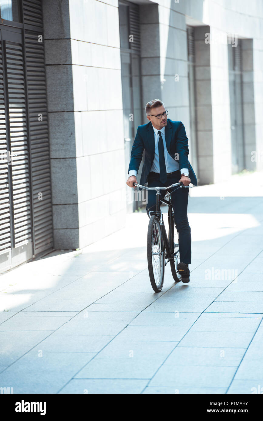 handsome middle aged businessman in suit and eyeglasses riding bicycle ...