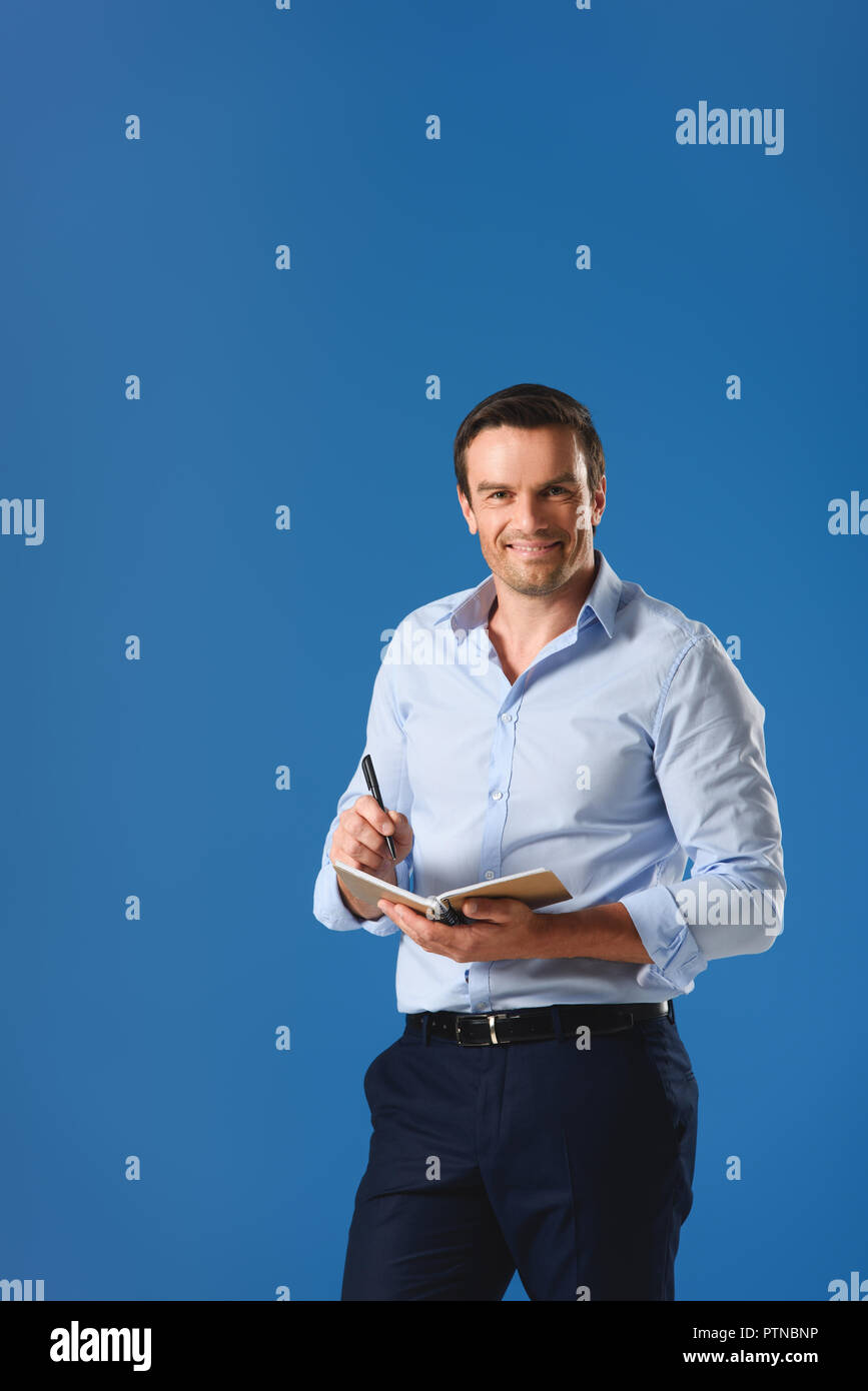 handsome businessman writing in notebook and smiling at camera isolated ...