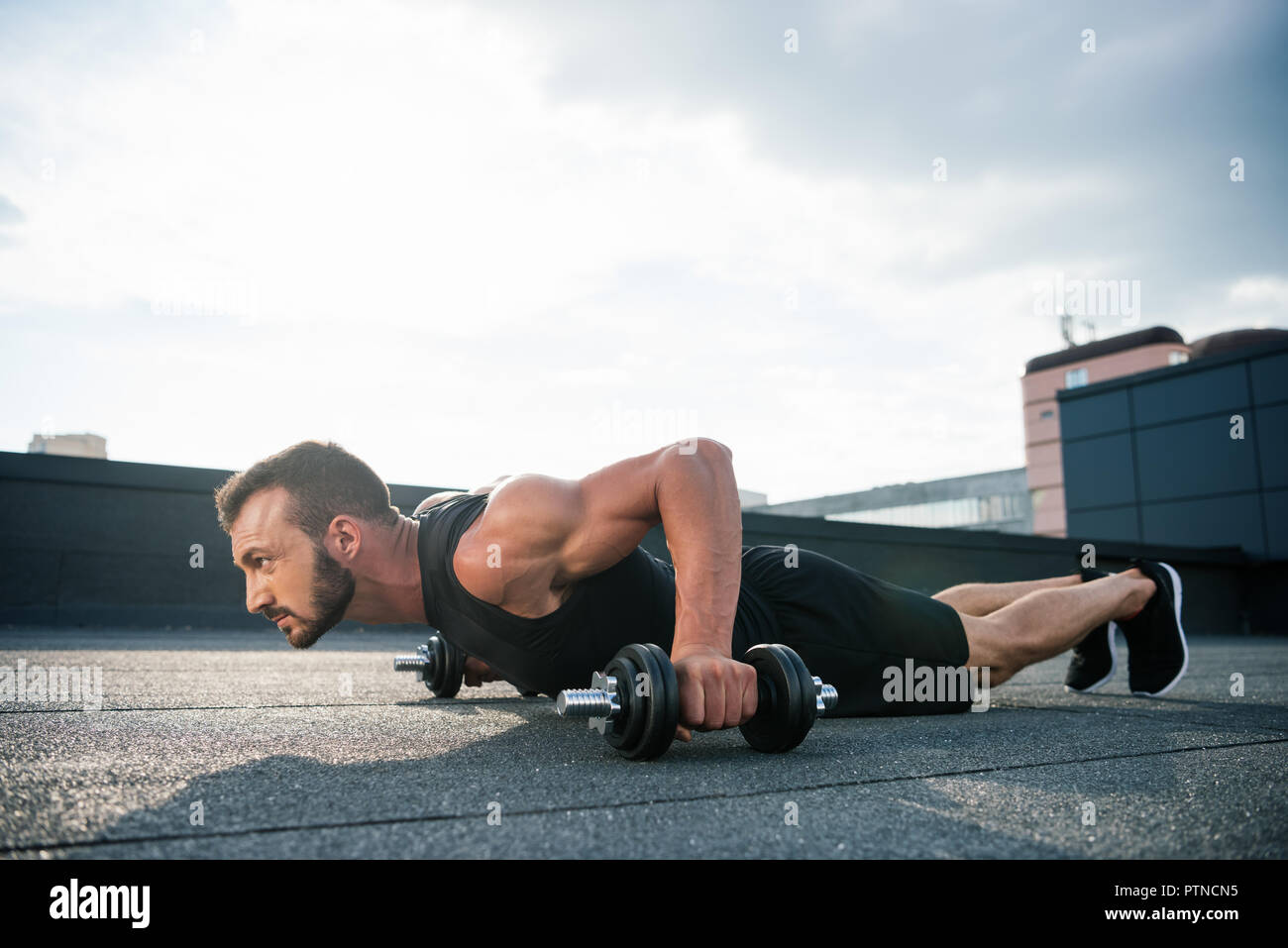 side view of handsome sportsman doing push ups with dumbbells on roof ...