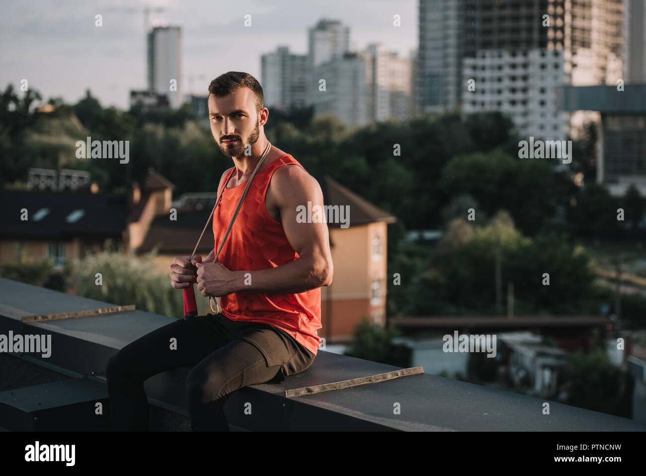 handsome sportsman with jumping rope on neck sitting on roof and ...