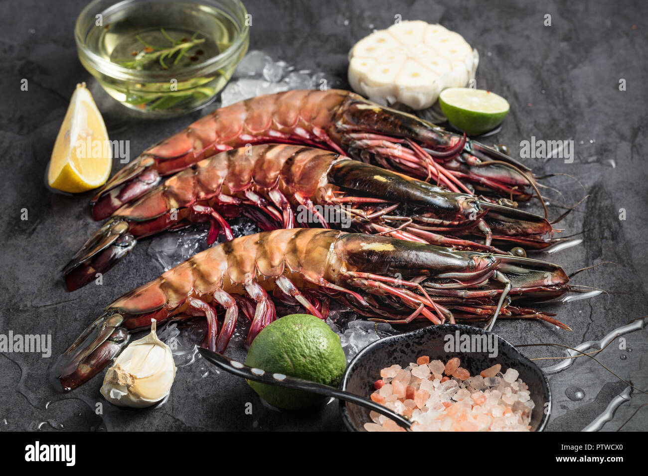 Three raw jumbo shrimps on ice with basic ingredients to cook on dark ...