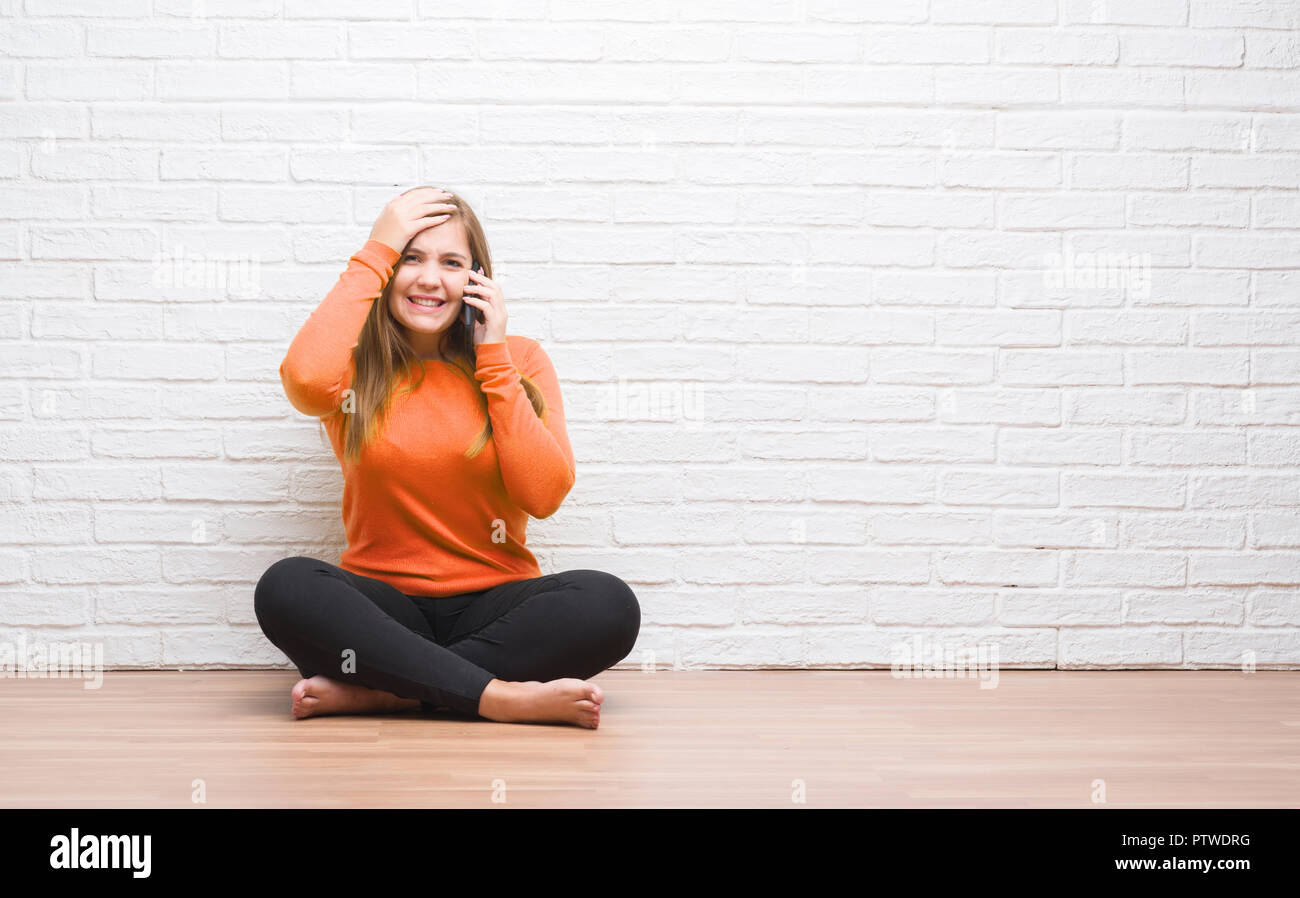 Young adult woman sitting on the floor over white brick wall calling ...