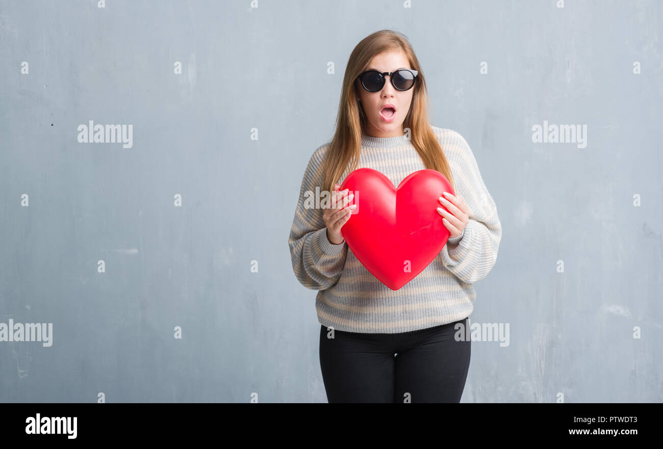 Young adult woman over grey grunge wall holding red heart in love ...
