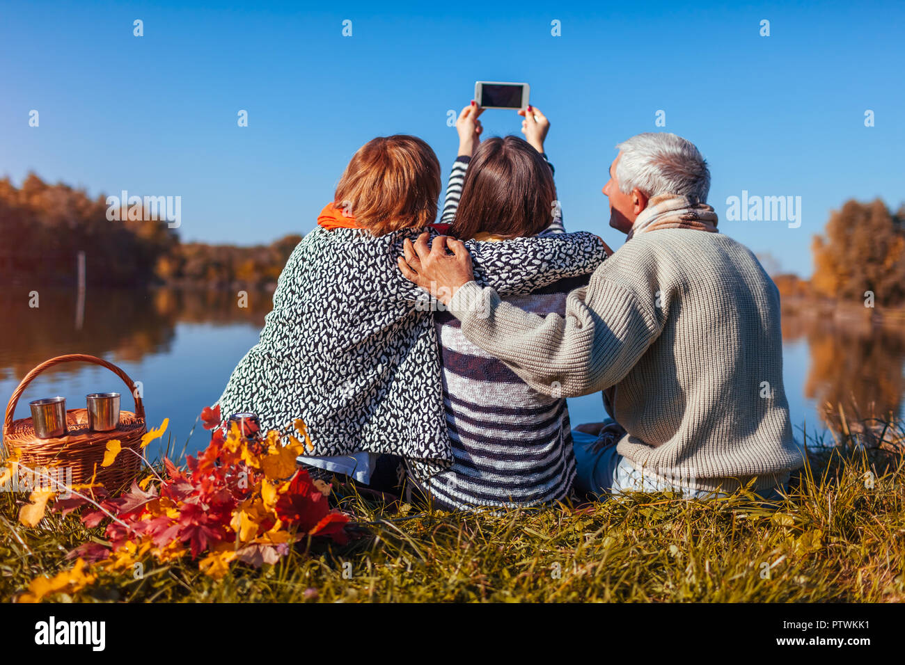 Senior parents taking selfie on phone by autumn lake with their adult ...