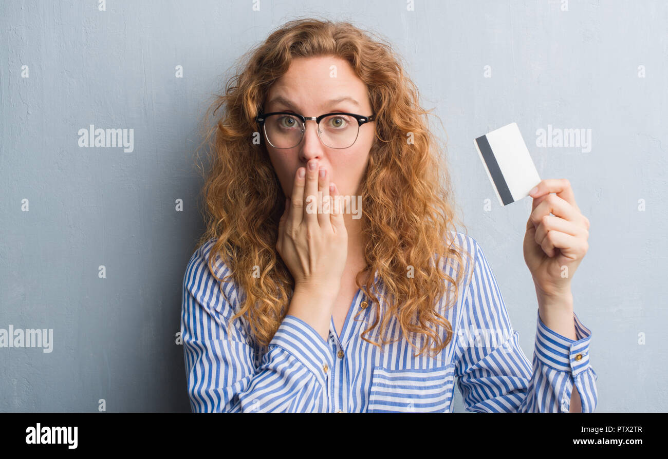 Young redhead woman over grey grunge wall holding credit card cover ...