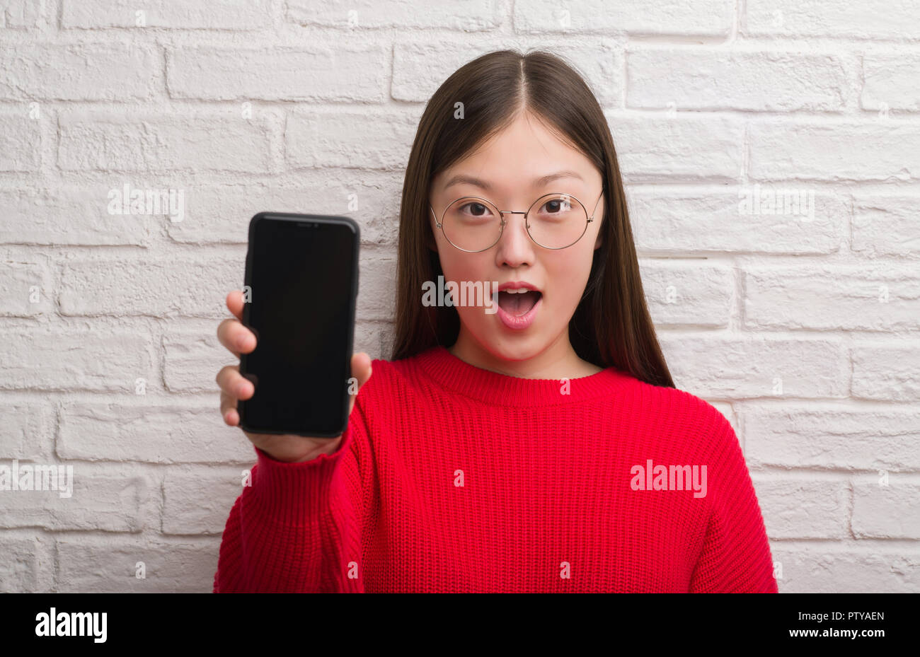 Young Chinese woman over brick wall showing smartphone scared in shock ...