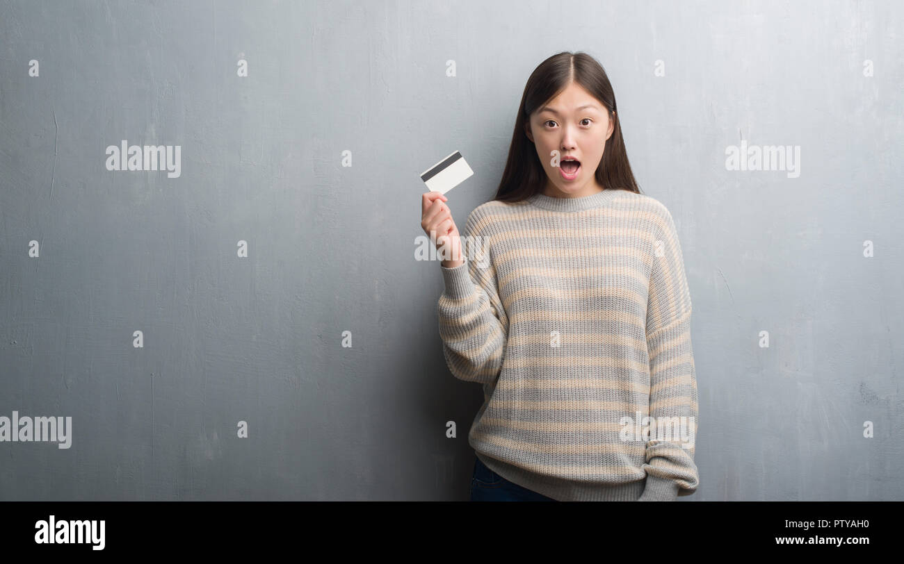 Young Chinese woman over grey wall holding credit card scared in shock ...