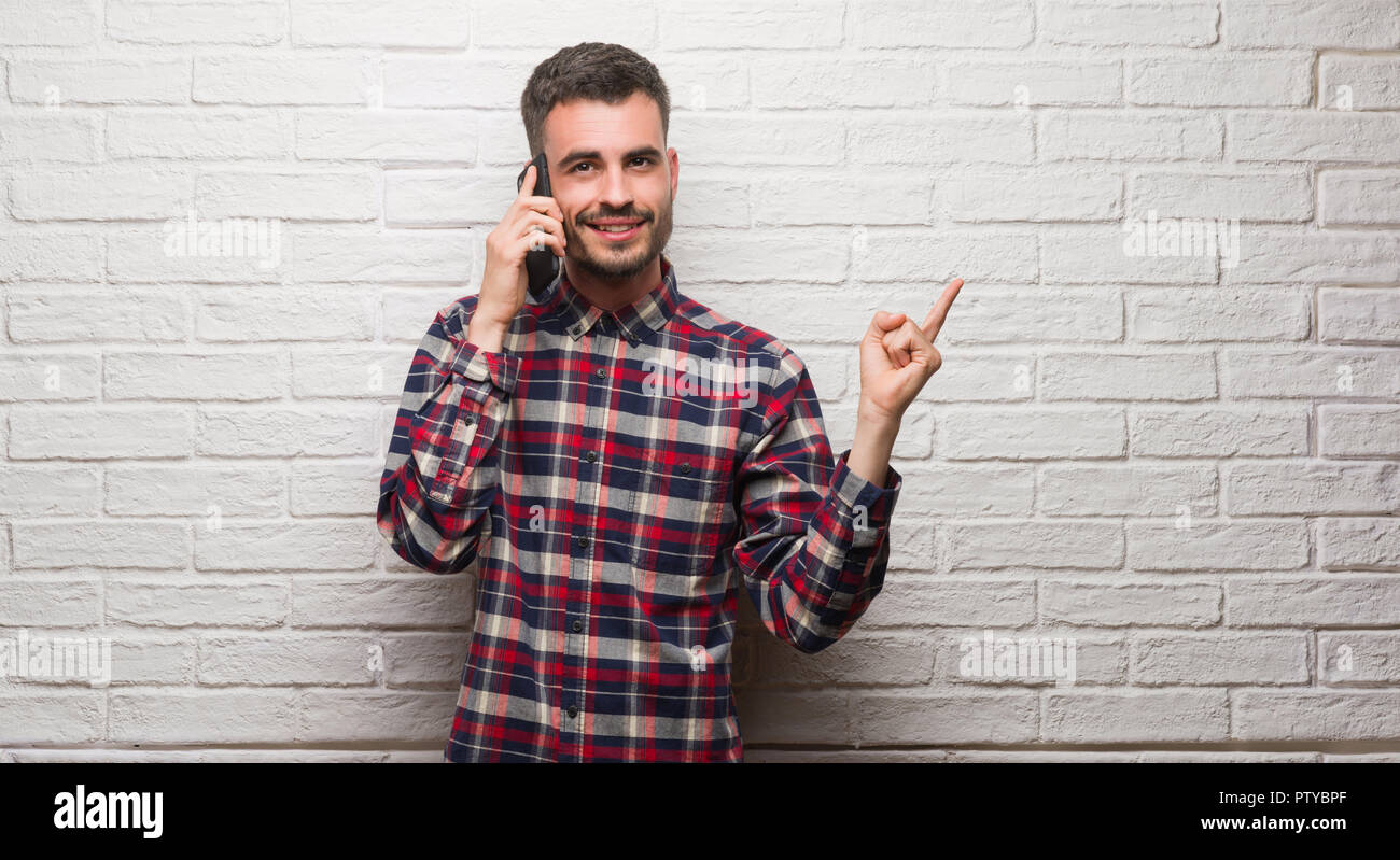 Young adult man talking on the phone standing over white brick wall ...