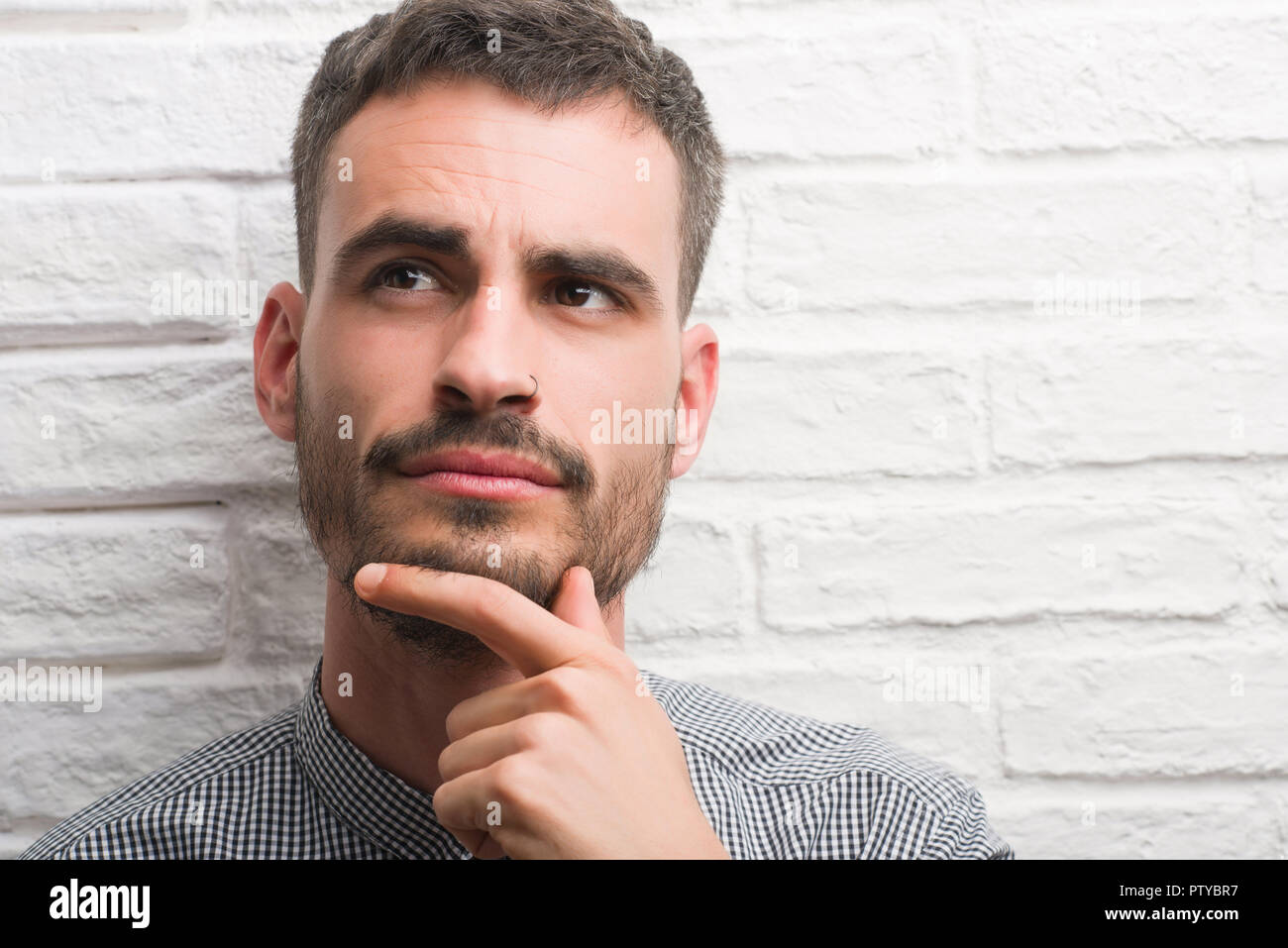 Young adult man standing over white brick wall serious face thinking ...