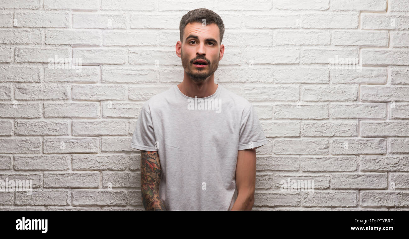 Young adult man standing over white brick wall scared in shock with a ...
