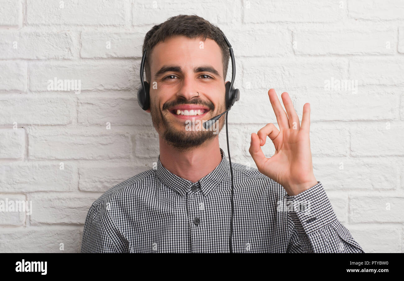 Young adult man over brick wall working as operator doing ok sign with ...
