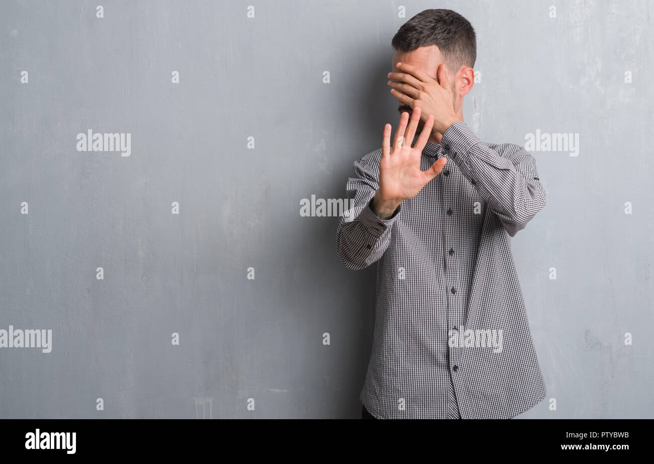 Young adult man standing over grey grunge wall covering eyes with hands ...