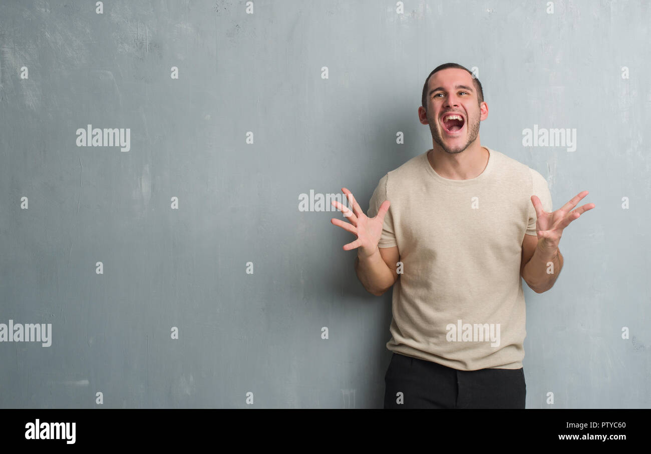 Young caucasian man over grey grunge wall crazy and mad shouting and ...