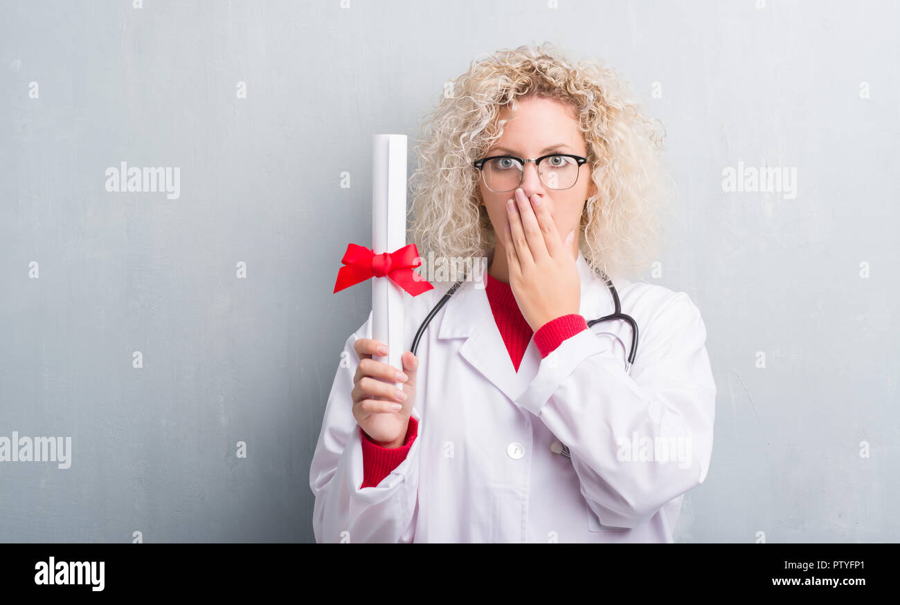 Young blonde doctor woman over grunge grey wall holding degree cover ...
