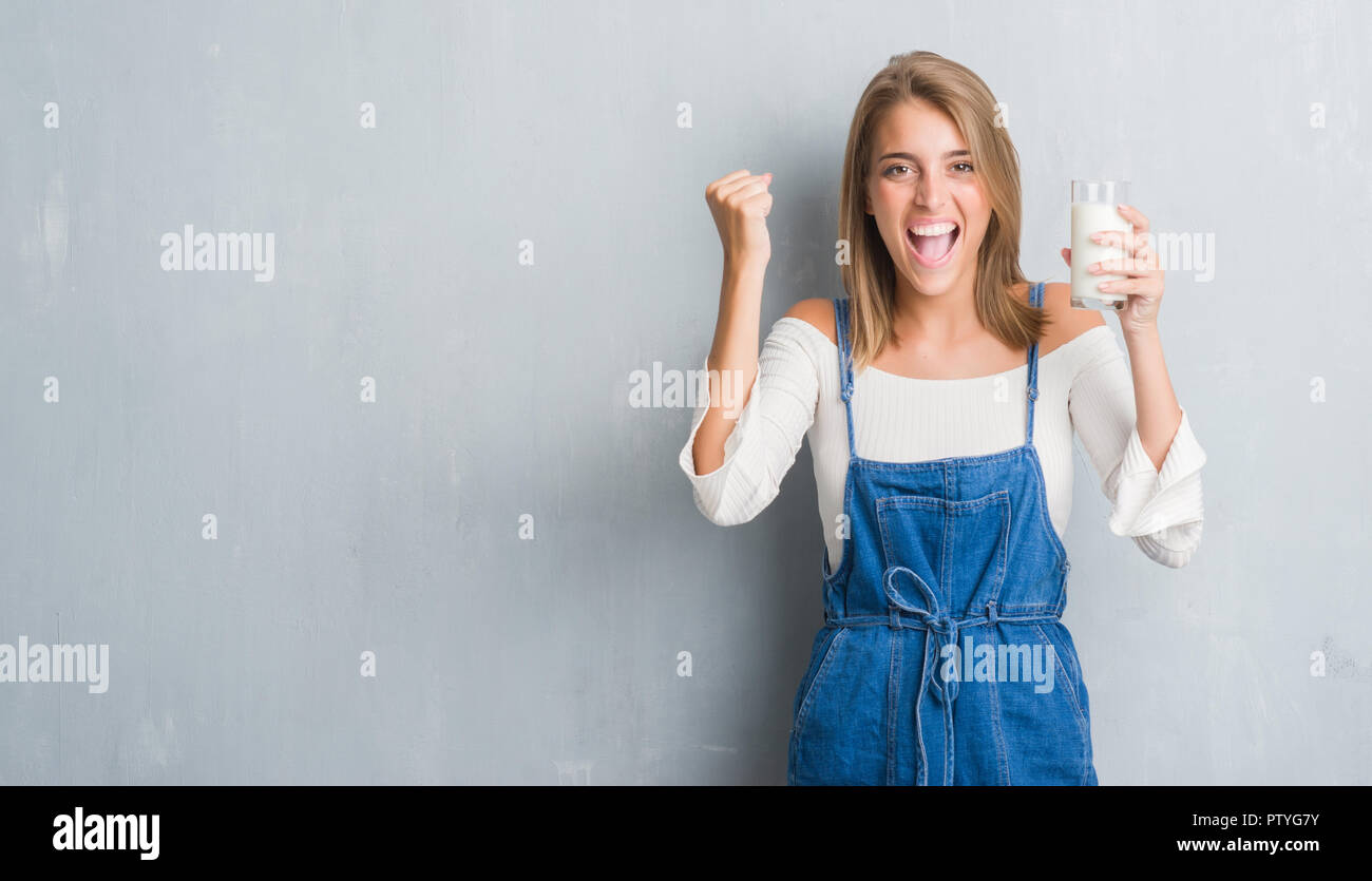 Beautiful young woman over grunge grey wall driking a glass of milk ...