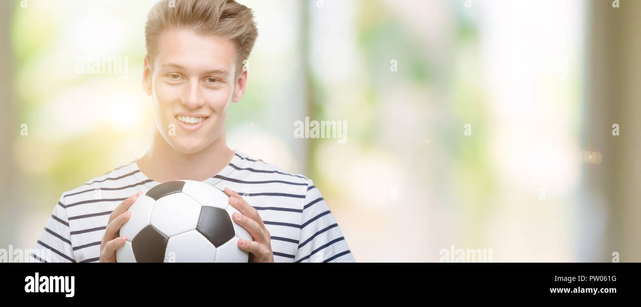 Young handsome blond man holding soccer ball with a happy face standing ...