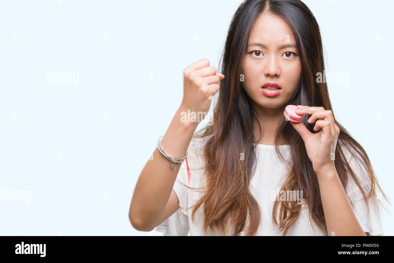 Young asian woman eating pink macaron sweet over isolated background ...