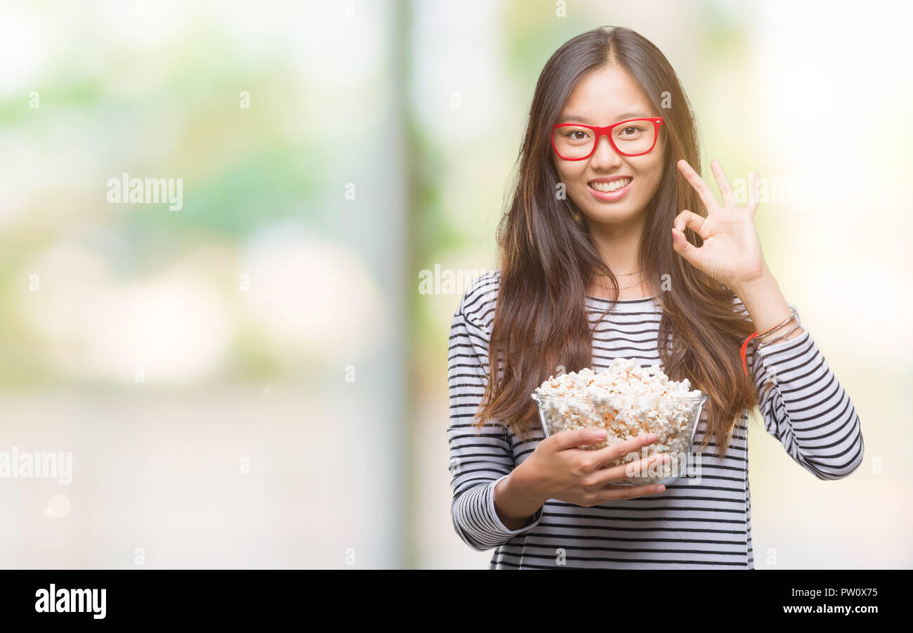 Young asian woman eating popcorn over isolated background doing ok sign ...