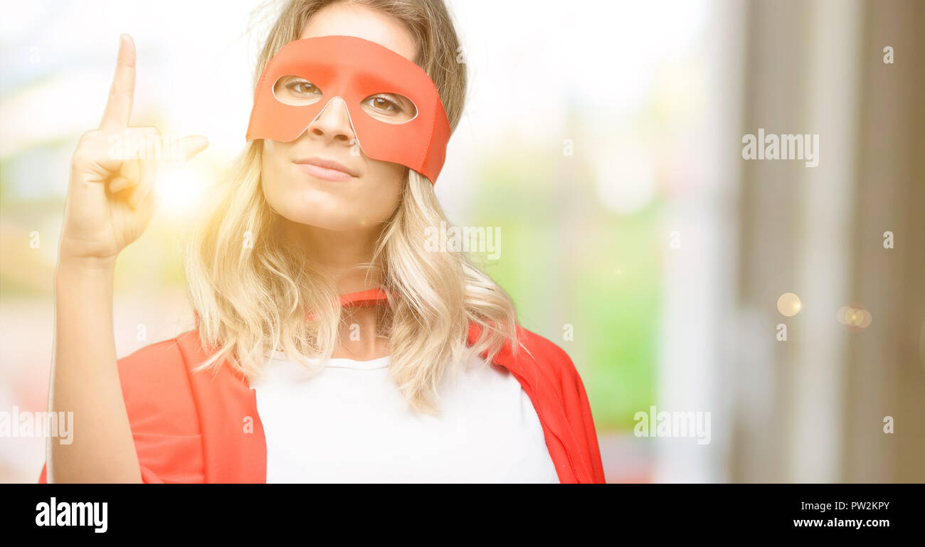 Young super hero woman wearing cape raising finger, the number one ...