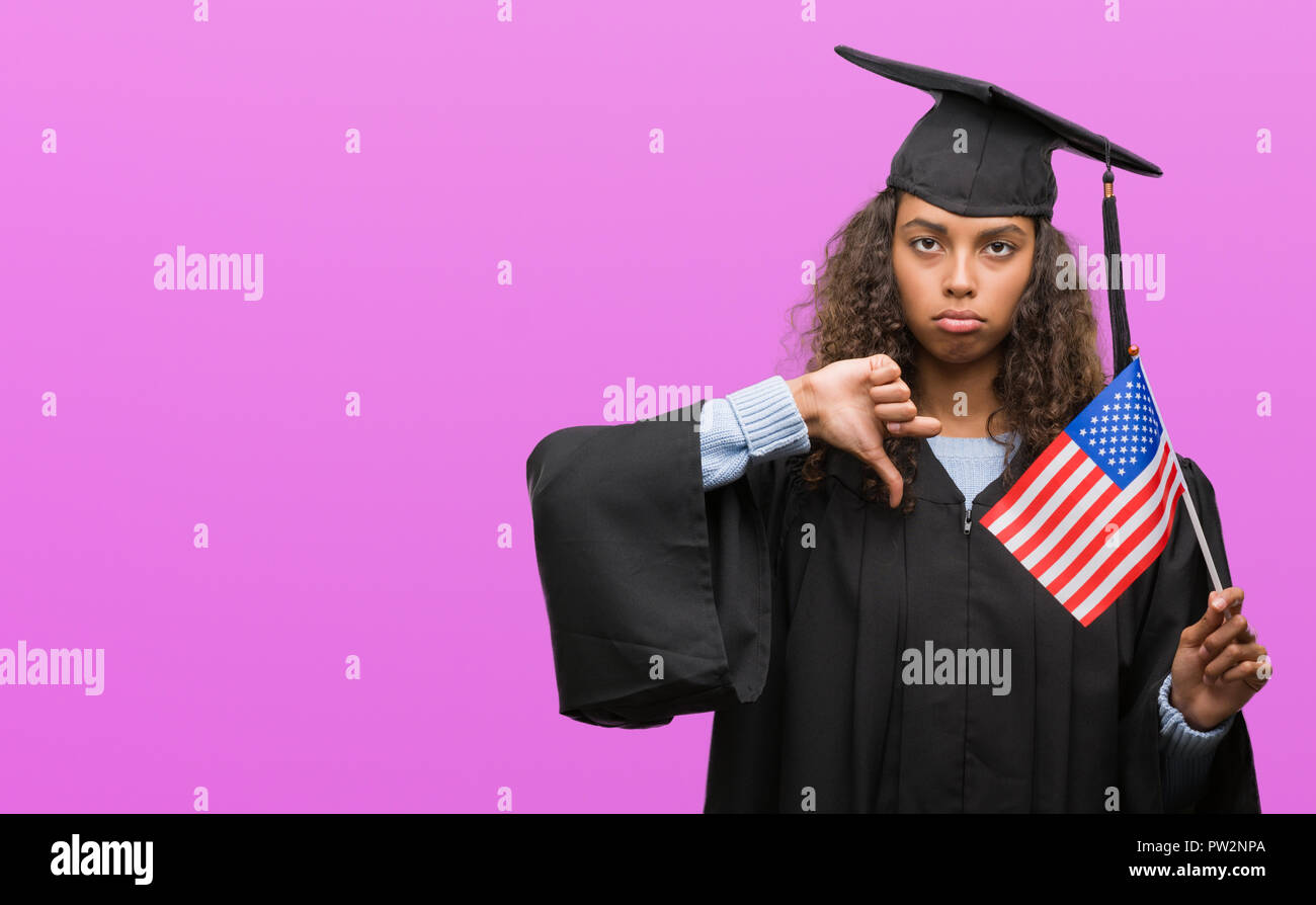 Young hispanic woman wearing graduation uniform holding flag of United ...