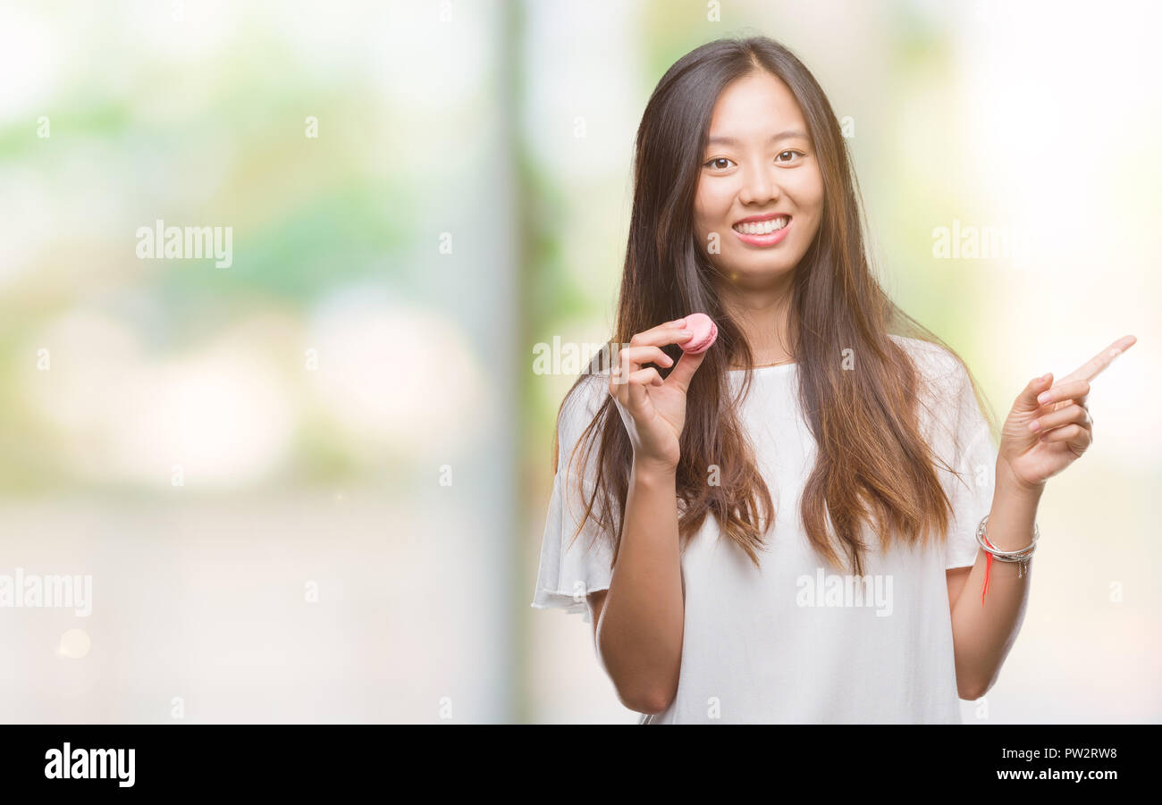 Young asian woman eating pink macaron sweet over isolated background ...