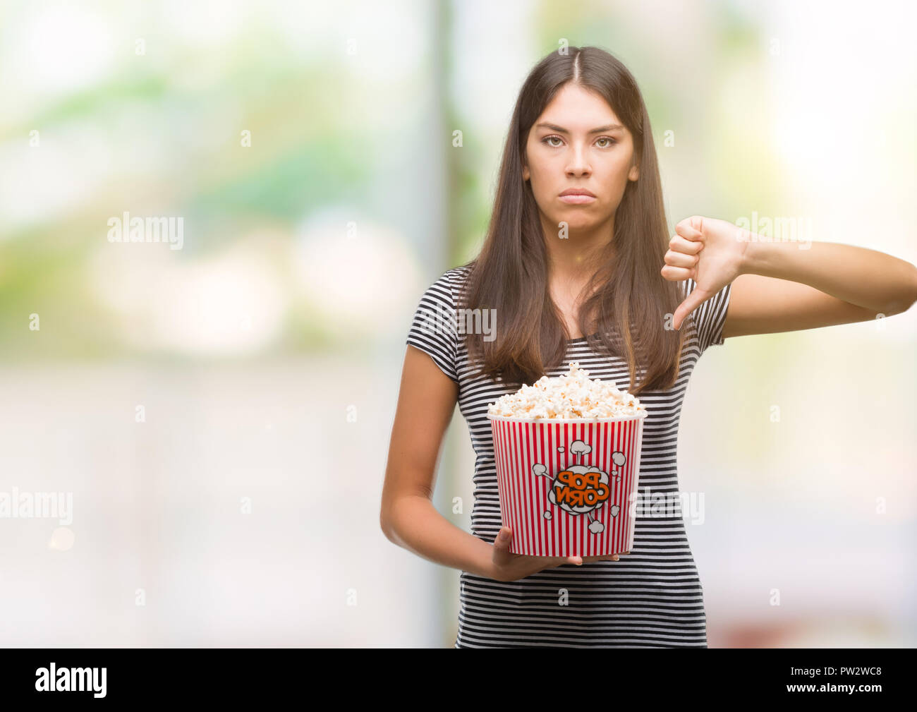 Young beautiful hispanic eating popcorn with angry face, negative sign ...