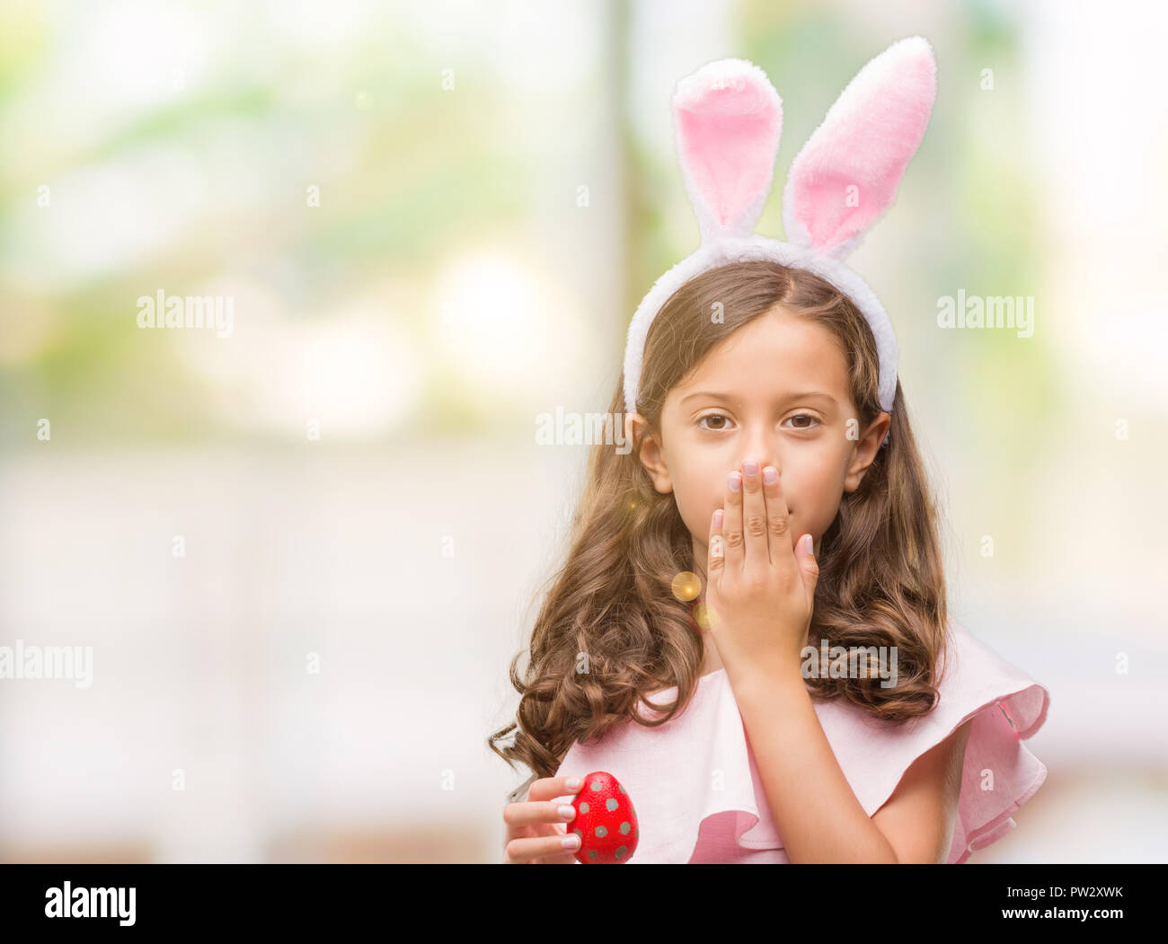 Brunette hispanic girl wearing easter rabbit ears cover mouth with hand ...