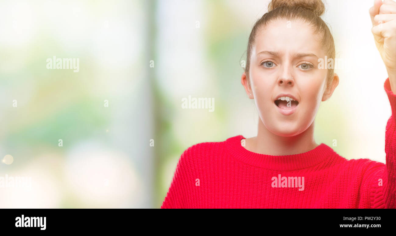 Young blonde woman wearing bun and red sweater angry and mad raising ...