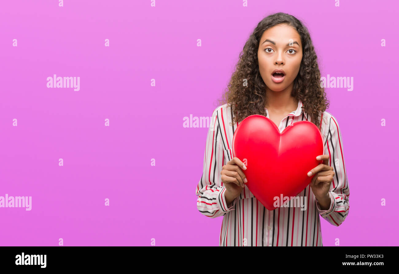 Young hispanic woman in love holding red heart scared in shock with a ...