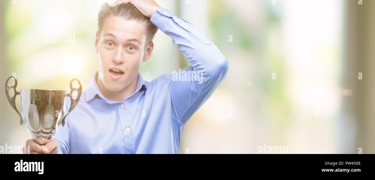 Young handsome blond man holding a trophy stressed with hand on head ...