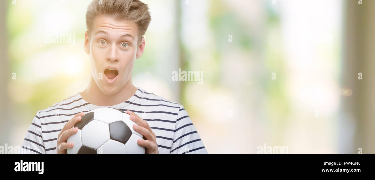 Young handsome blond man holding soccer ball scared in shock with a ...