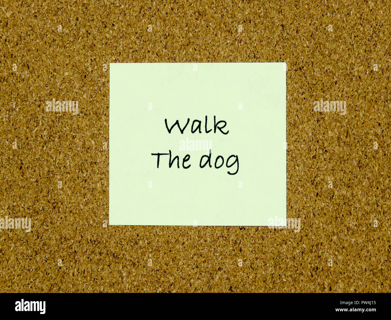 A yellow sticky note with walk the dog written on it on a cork board ...