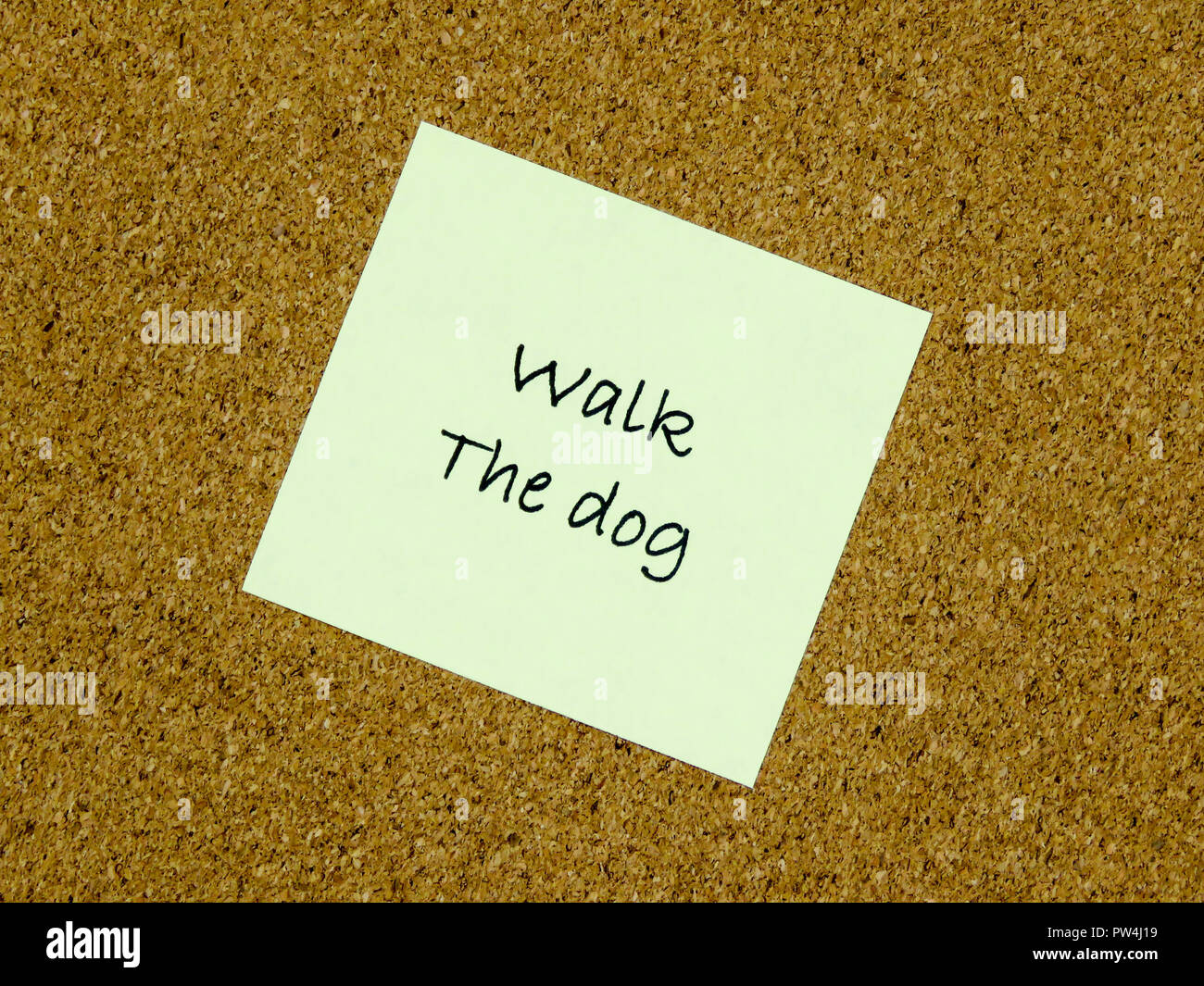 A yellow sticky note with walk the dog written on it on a cork board ...