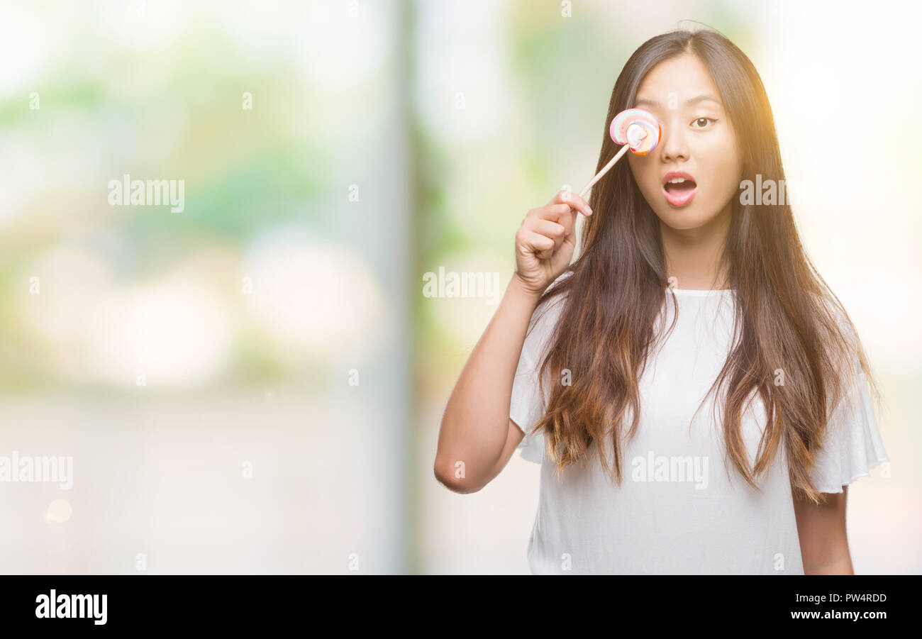 Young asian woman eating lollipop candy over isolated background scared ...