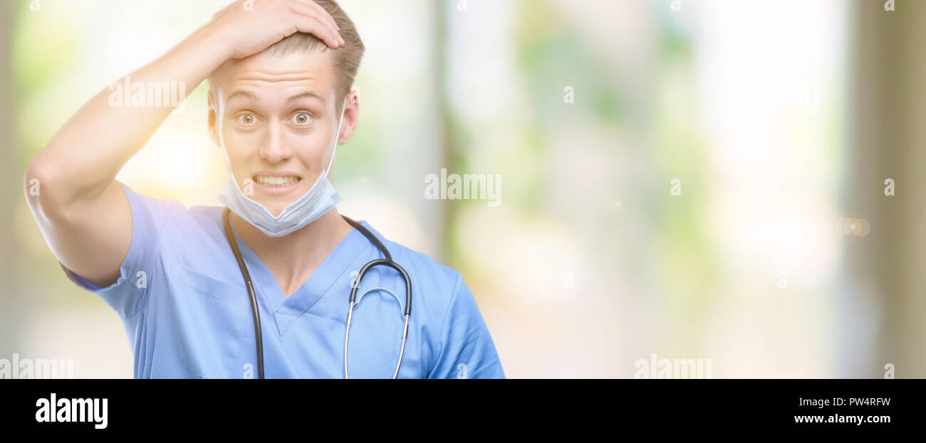 Young handsome blond doctor stressed with hand on head, shocked with ...