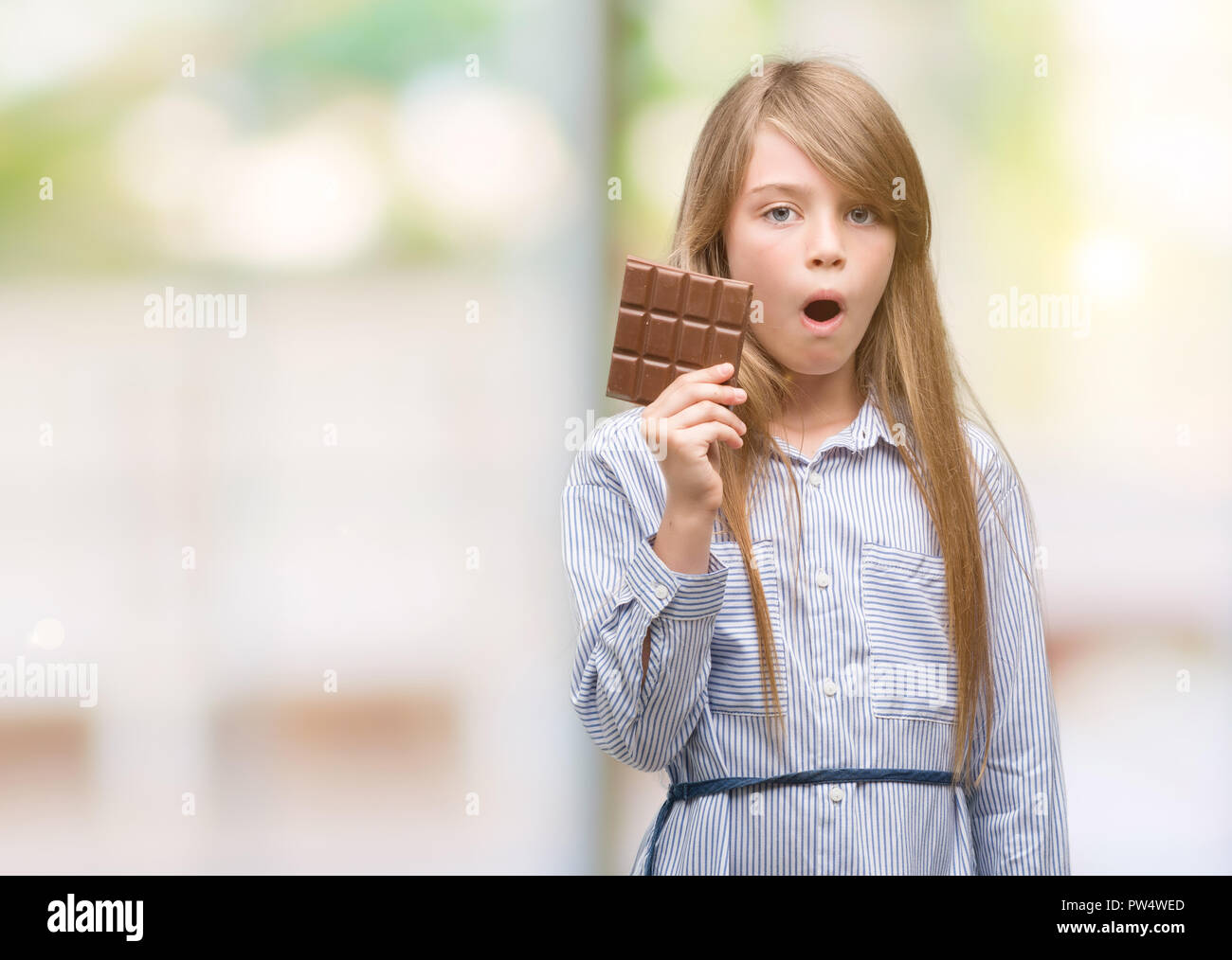 Young blonde child holding chocolate bar scared in shock with a ...