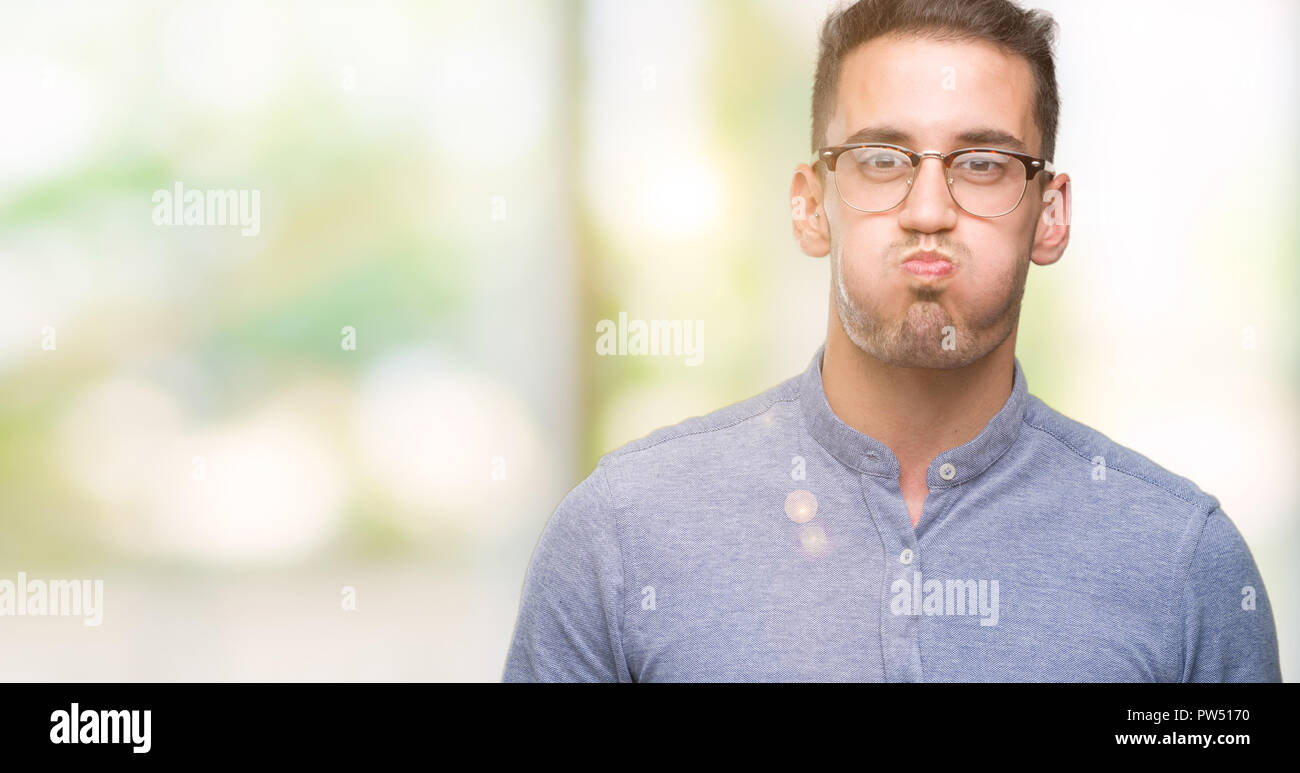 Handsome young elegant man wearing glasses puffing cheeks with funny ...