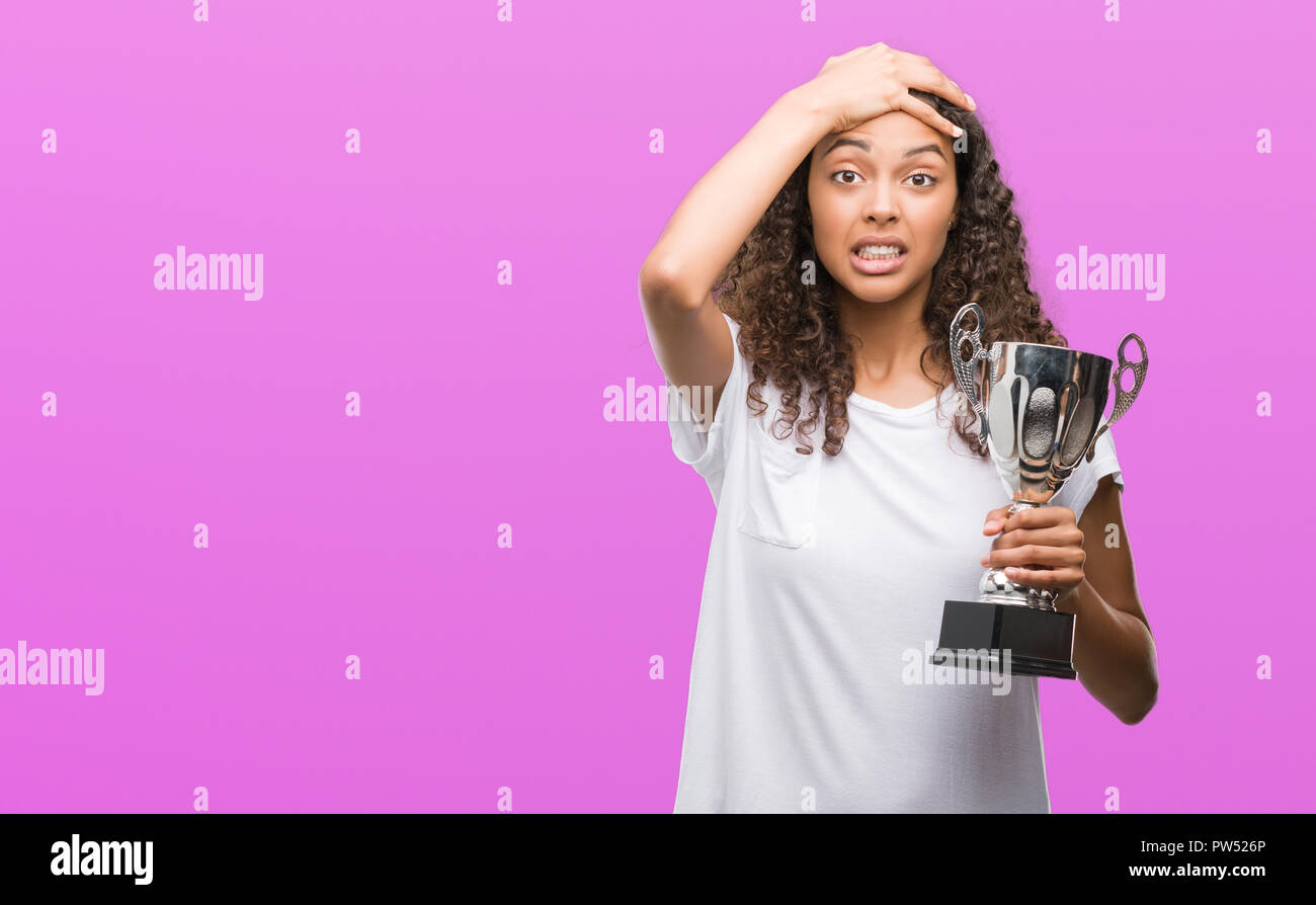 Young hispanic woman holding trophy stressed with hand on head, shocked ...