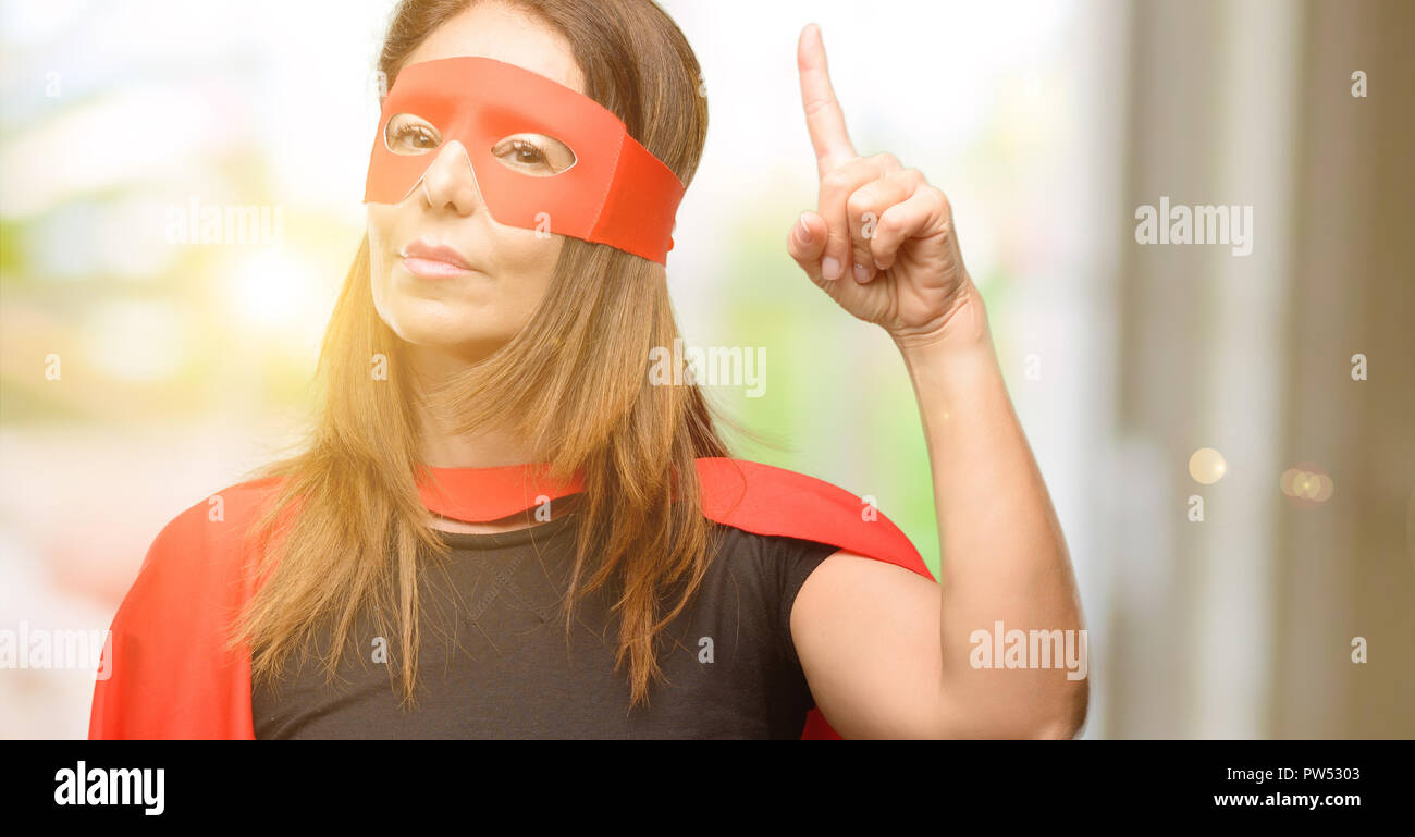 Middle age super hero woman wearing red mask and cape raising finger ...